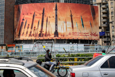 A motorcyclist speaks on a phone on April 15, 2024, while driving past a billboard depicting named Iranian ballistic missiles in service, with text in Arabic reading “the true promise“ and in Farsi “Israel is weaker than a spider's web“, in Valiasr Square in central Tehran.